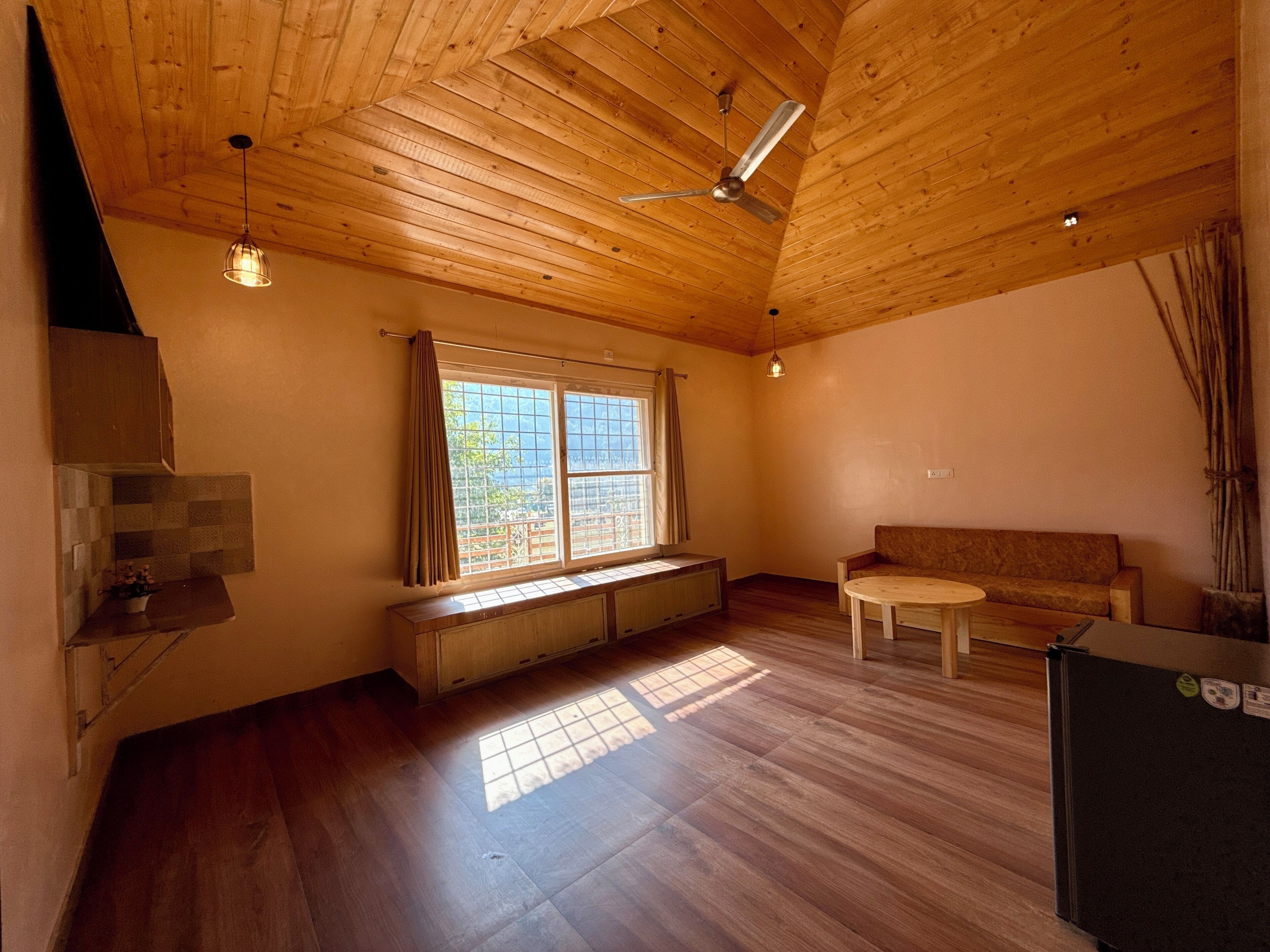 Quarto com piso de madeira, ventilador de teto, janela com cortinas, sofá e uma mesa.
