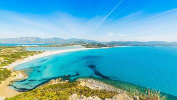 Paisagem costeira com águas turquesa cristalinas, uma praia arenosa e montanhas ao longe.