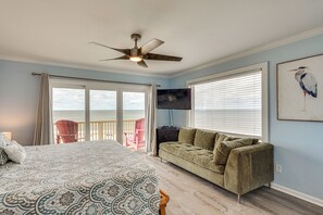 Quarto com cama, sofá verde, ventoinha no teto, televisão e varanda com vista para o mar.