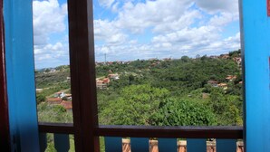 Vista da janela com molduras azuis, mostrando um morro com casas e vegetação.
