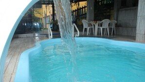 Piscina com um elemento aquático, rodeada por um deck de madeira e cadeiras de plástico branco.