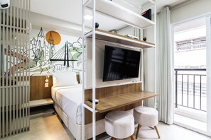 Quarto moderno com cama, TV fixada numa prateleira de madeira, escrivaninha e varanda com guarda-corpo.