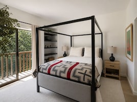 Uma cama de dossel com colcha estampada em padrão geométrico, mesas de cabeceira com luminárias e vista para uma varanda com uma planta.