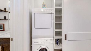 Uma lavanderia com eletrodomésticos empilhados, incluindo uma secadora e uma máquina de lavar, e uma prateleira com uma garrafa por cima.