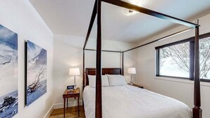 Uma cama com dossel e colunas, uma mesinha de cabeceira com abajur, e uma janela com vista para árvores.