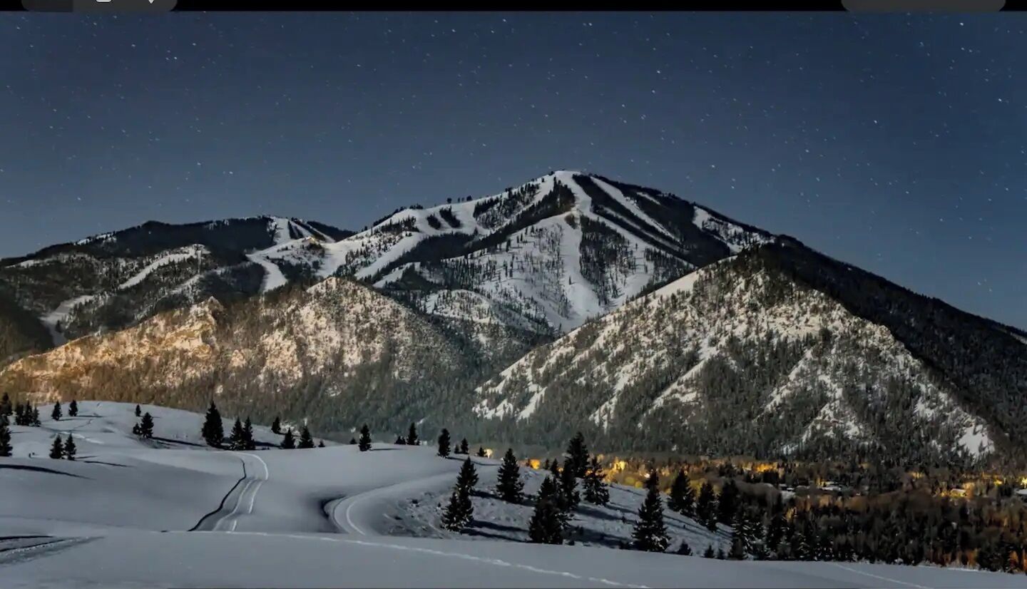 Paisagem montanhosa coberta de neve à noite, com uma estrada sinuosa e edifícios iluminados no vale.