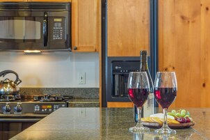 Uma cozinha com micro-ondas, fogão e um armário de madeira. Há duas taças de vinho tinto no balcão, uma garrafa de vinho e um prato com uvas e bolachas.