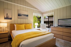 Quarto de hotel com uma cama grande, televisão, escrivaninha com abajur e janela com vista para árvores.