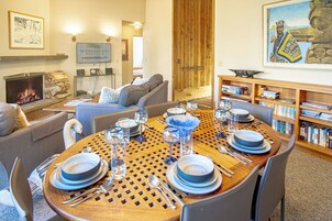 Uma área de refeições com mesa de madeira posta para quatro pessoas, cercada por estantes de livros e lareira.