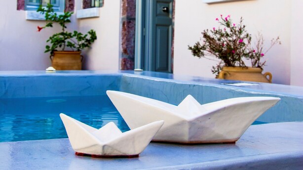Uma área à beira da piscina com uma piscina azul, um barquinho de papel branco, e um edifício com porta e janelas azuis.