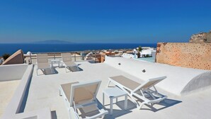 Terraço no telhado com cadeiras e mesas de estar brancas, com vista para o mar e montanhas ao longe.