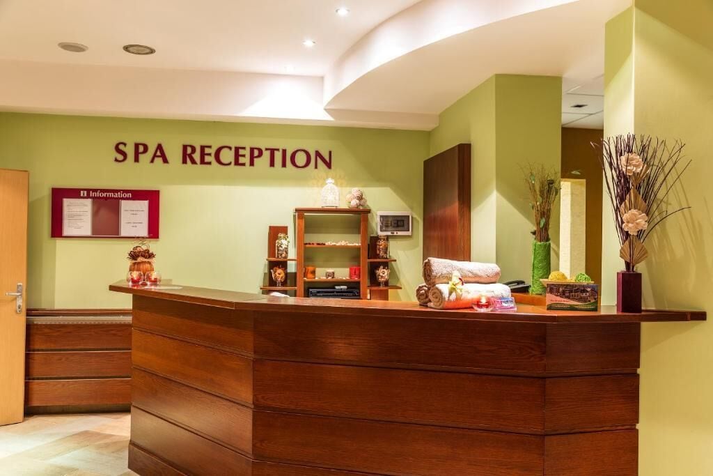 Uma área de recepção de spa com um balcão de madeira, uma placa 'SPA RECEPTION', um pequeno quadro de informações e objetos decorativos como um vaso e um relógio.