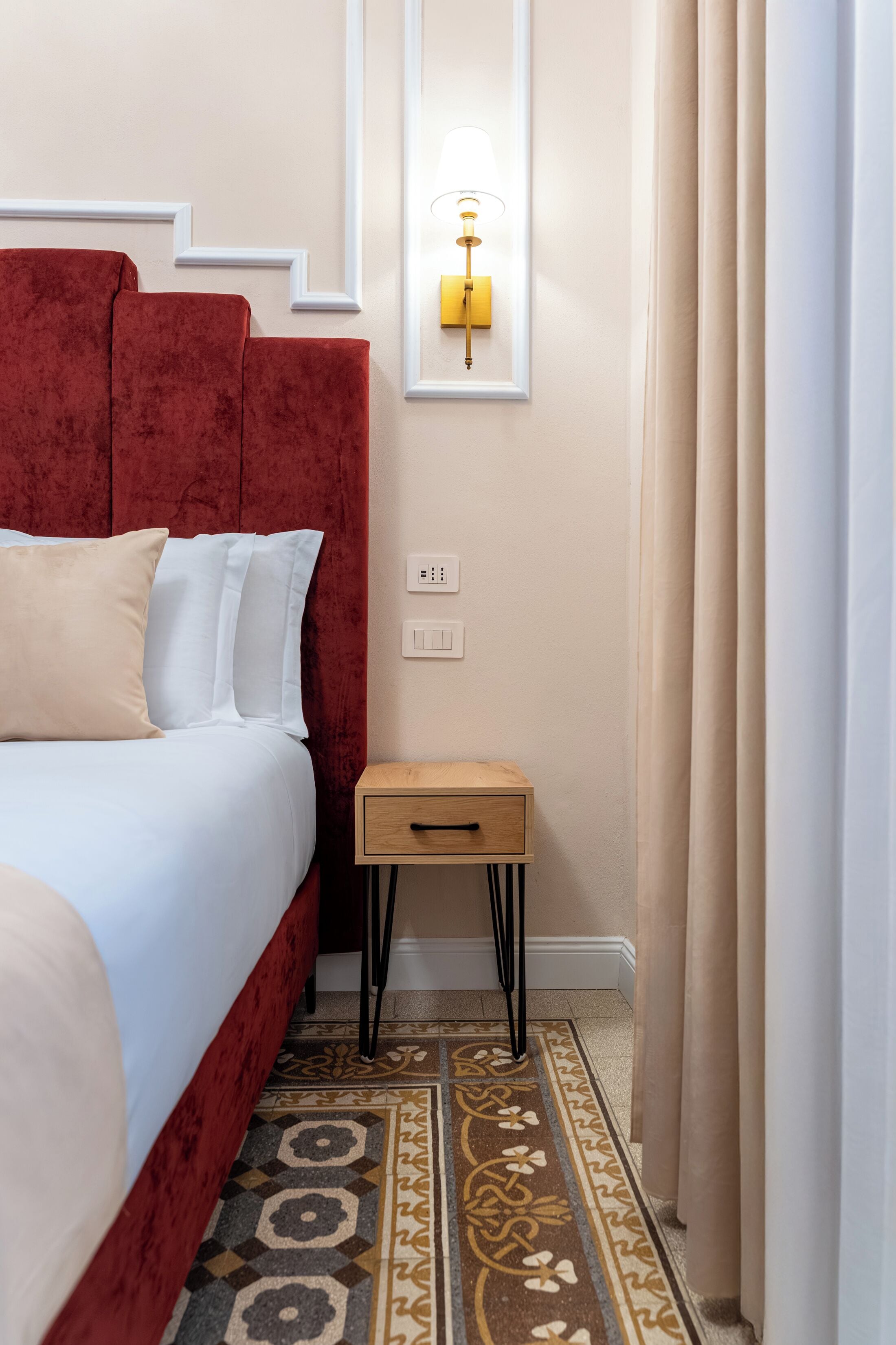 Quarto de hotel com cabeceira vermelha, mesa de cabeceira com gaveta, tapete estampado e luminária fixada na parede.