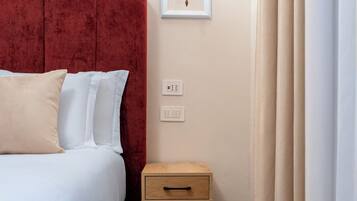 Quarto de hotel com cabeceira vermelha, mesa de cabeceira com gaveta, tapete estampado e luminária fixada na parede.