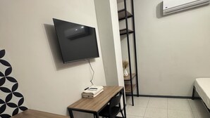 Quarto pequeno e moderno com escrivaninha de madeira, cadeira preta, televisão fixada na parede e ar condicionado branco.