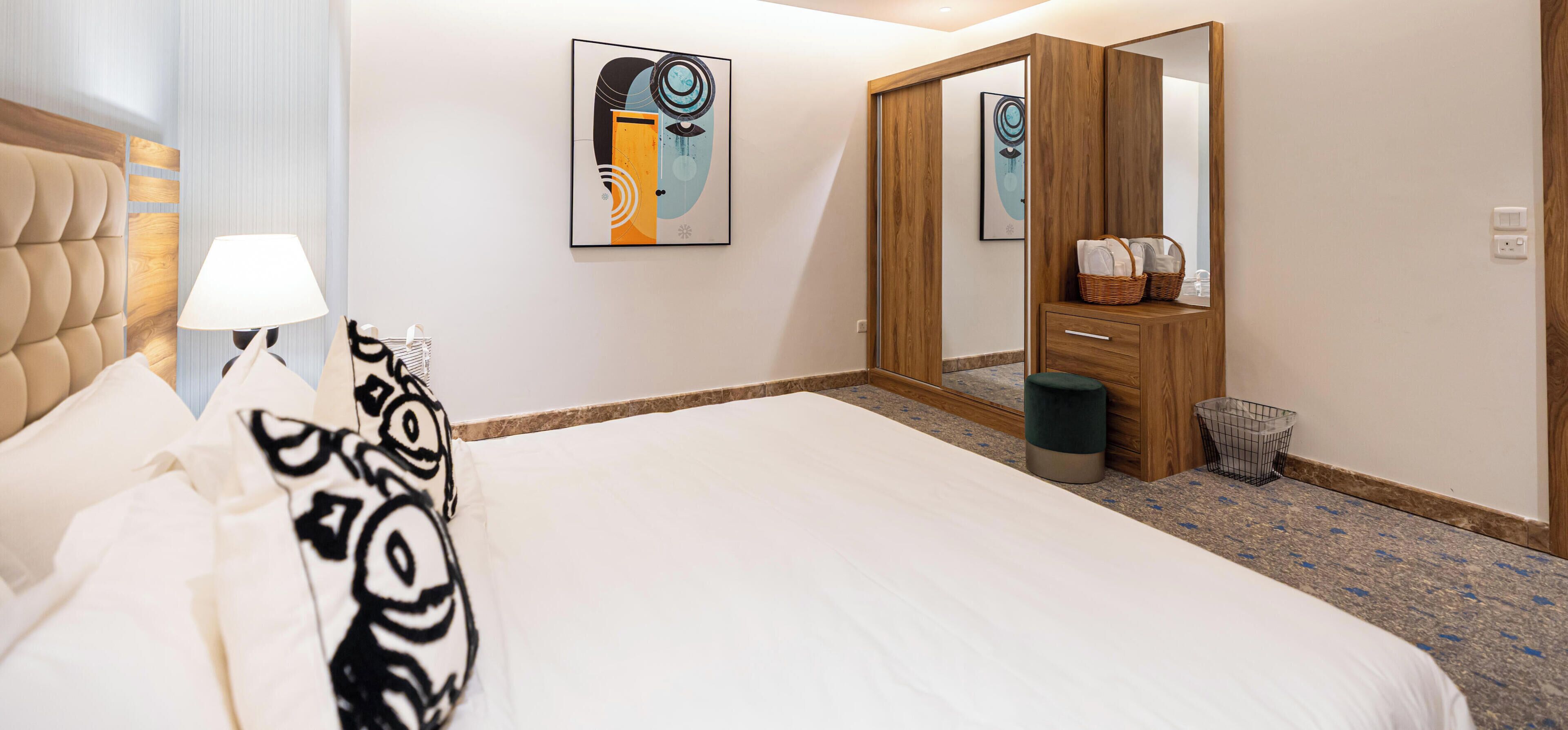 Quarto de hotel com uma cama grande, cabeceira de madeira, mesa de cabeceira com abajur, penteadeira com espelho e obras de arte abstratas nas paredes.