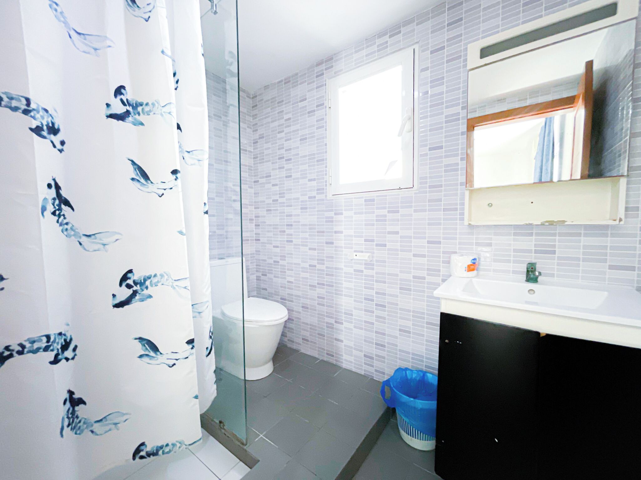 Uma casa de banho com cortina de chuveiro com padrão azul e branco, sanita branca, lavatório com espelho em cima e um caixote de lixo azul.