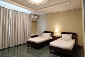 Quarto de hotel com duas camas de solteiro, mesa de cabeceira com luminária, janela com cortinas e ar condicionado na parede.