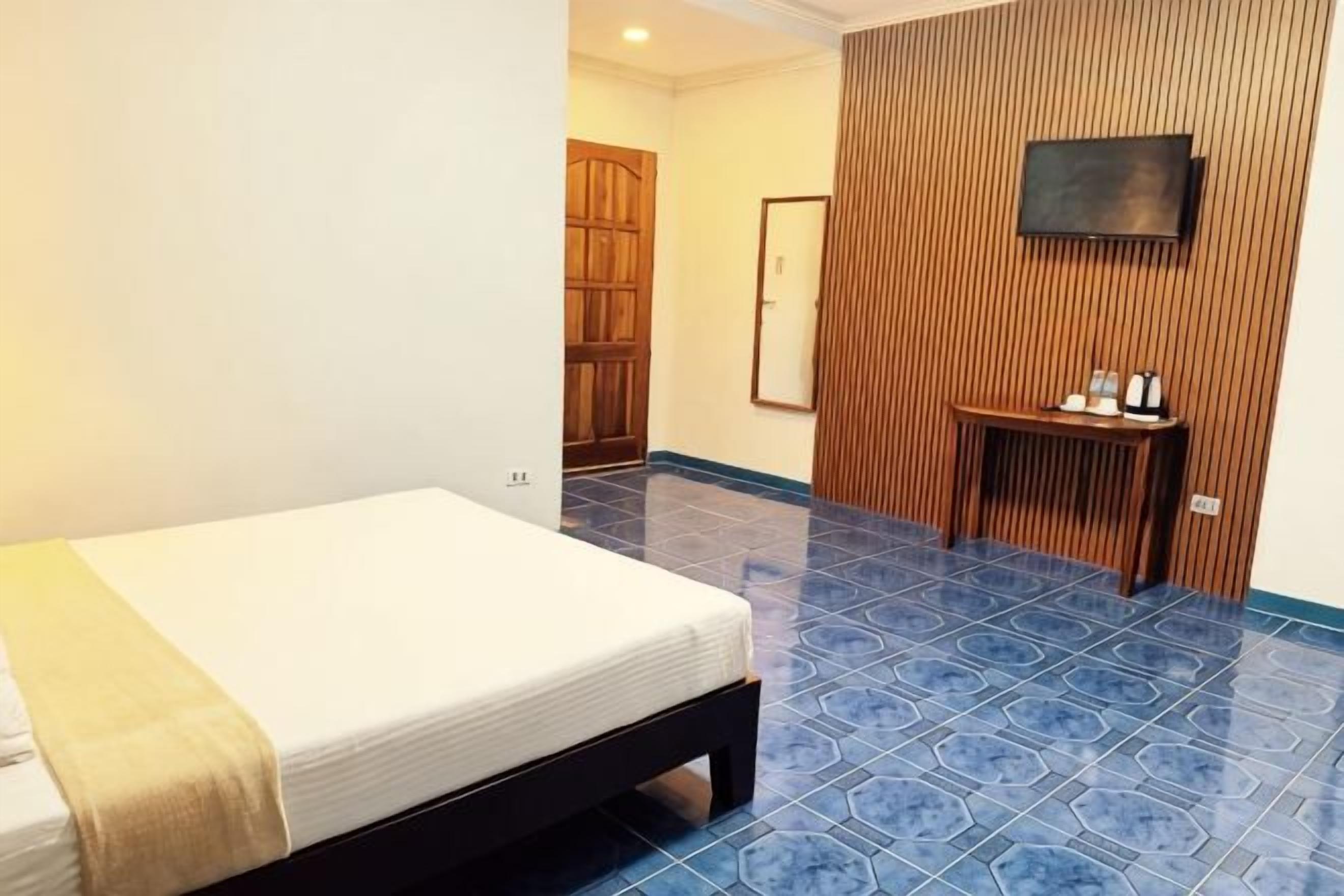 Quarto de hotel com cama, parede de painéis de madeira, televisão e uma pequena mesa com objetos.
