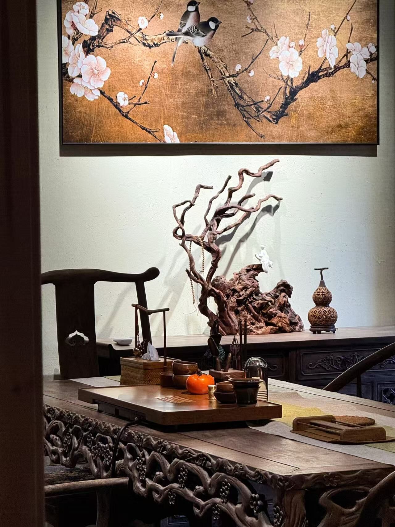 Um interior tradicional com um quadro de pássaros em um galho, uma cadeira de madeira e uma mesa com vários objetos.