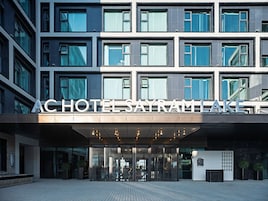 Uma entrada de hotel moderna com fachada de vidro transparente, uma placa com a inscrição 'ACHOTEL SAYRAMLAKE' e uma área de acesso pavimentada.