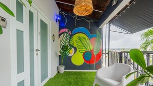Quarto com piso de grama sintética verde, uma cadeira branca, um mural colorido e uma varanda com vista.