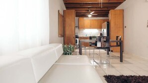Uma sala de estar moderna com um sofá branco, um tapete felpudo escuro, vigas de madeira no teto e uma cozinha ao fundo.