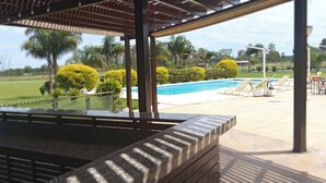Uma área à beira da piscina com um espaço de estar coberto e vista para a piscina e vegetação ao redor.