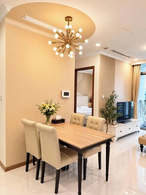 Quarto de hotel com área de jantar, apresentando uma mesa e cadeiras de madeira, um lustre moderno, uma televisão sobre um móvel e uma janela grande com cortinas.