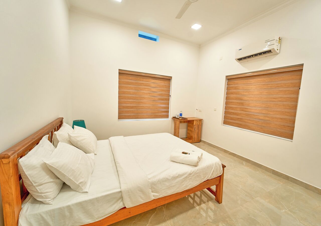 Quarto com cama de madeira, roupa de cama branca, mesa de cabeceira, dois aparelhos de ar condicionado fixados na parede e persianas de madeira nas janelas.