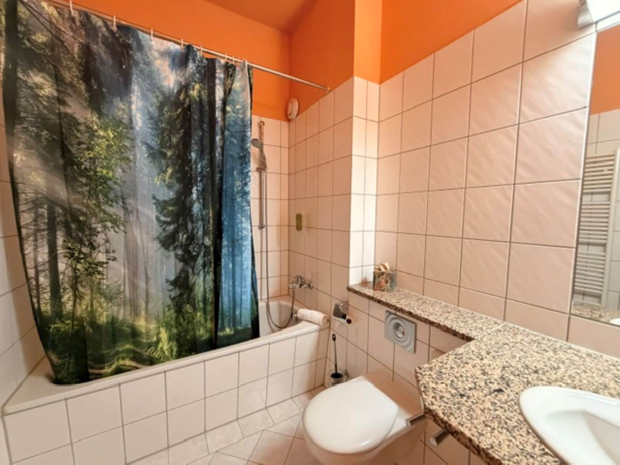 Casa de banho com cortina de chuveiro com cena de floresta, sanita e lavatório com bancada.