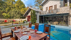 Mesa de jantar posta para o café da manhã, vista para a piscina, arquitetura de casa de pedra e vegetação exuberante.