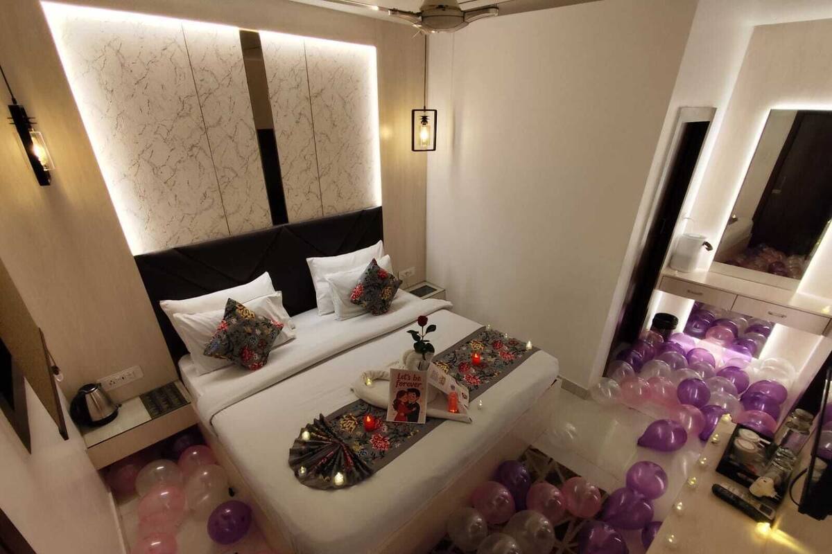 Quarto de hotel com uma cama grande, uma mesinha de cabeceira e um criado-mudo. Há balões no chão e uma pequena mesa com uma vela e um cartão.