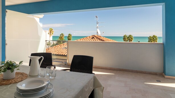 Varanda com mesa posta para refeições, vista para uma praia com palmeiras e céu claro.