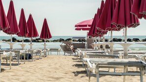Uma praia com fileiras de cadeiras de banho brancas e guarda-sóis vermelhos, com o oceano e a costa rochosa ao fundo.