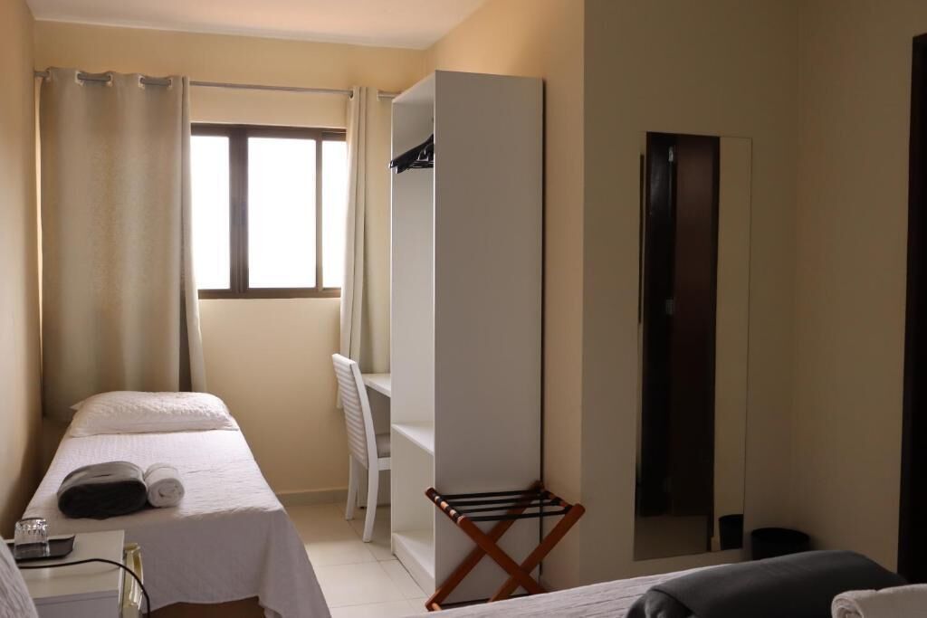 Quarto de hotel com duas camas de solteiro, um guarda-roupa, um espelho e uma janela com cortinas.