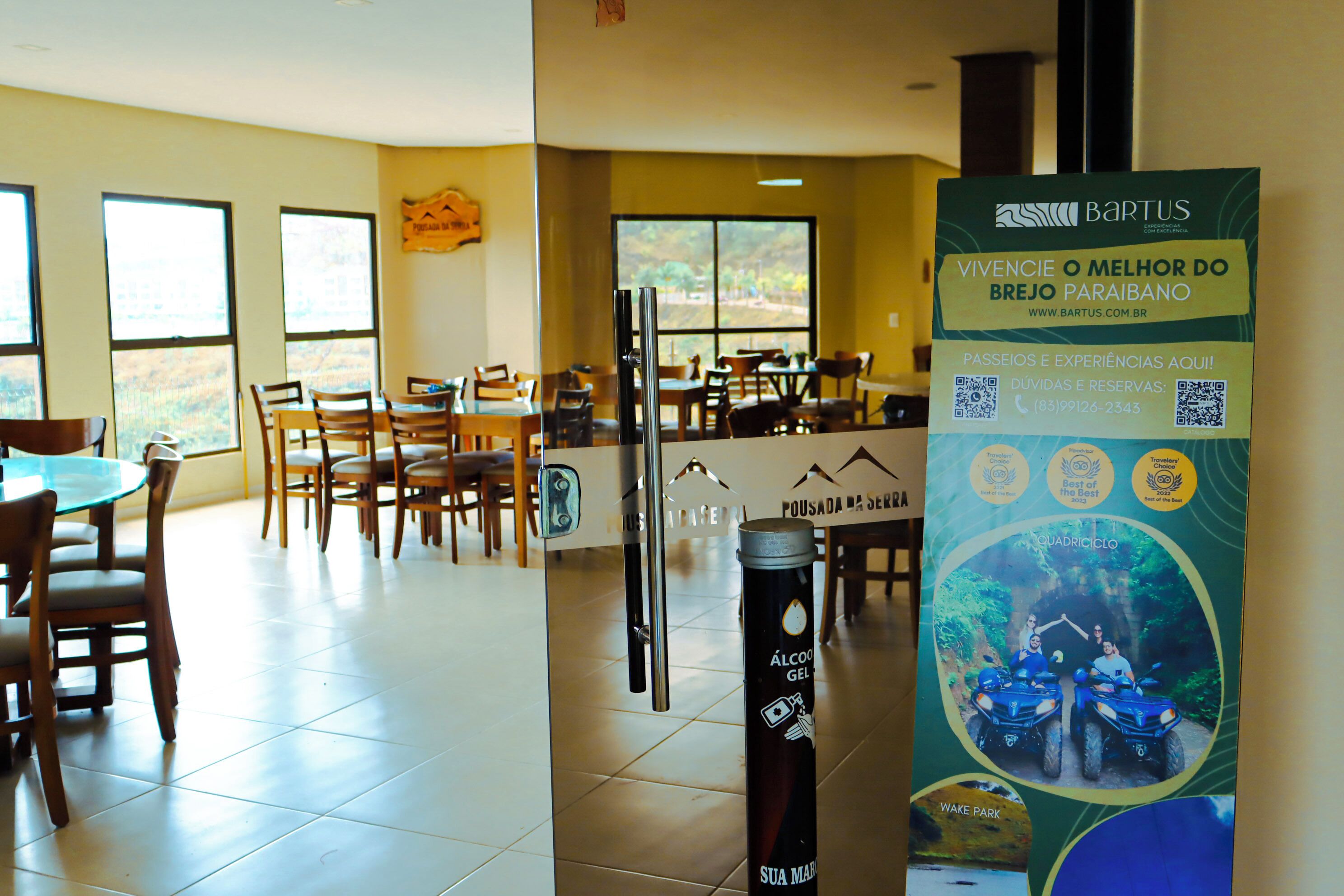 Restaurante com mesas e cadeiras de madeira, uma mesa com tampo de vidro e um anúncio de experiência de parapente.