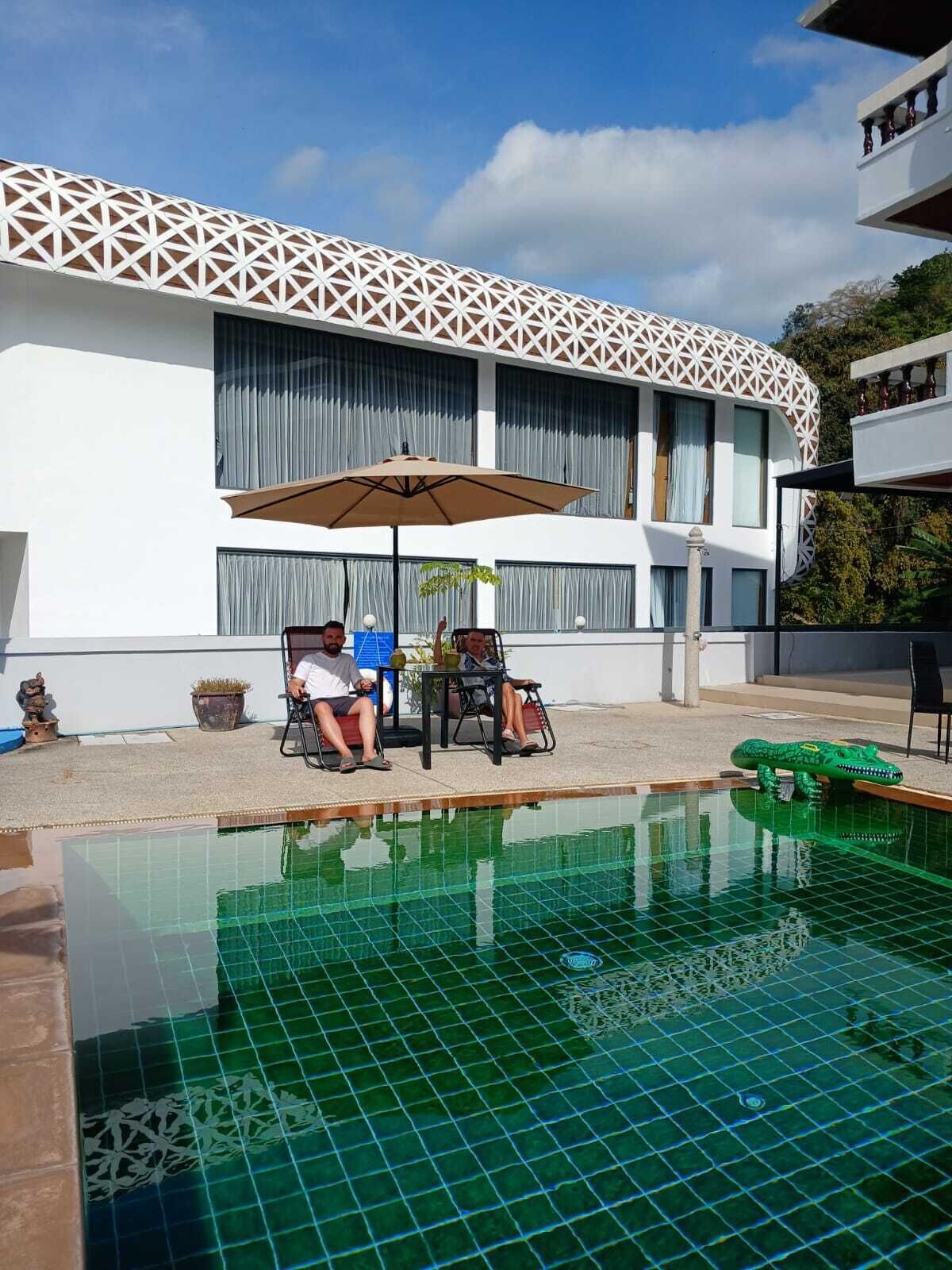 Uma área de piscina com duas pessoas sentadas sob um grande guarda-sol, um crocodilo insuflável verde e um edifício com fachada branca e telhado estampado.
