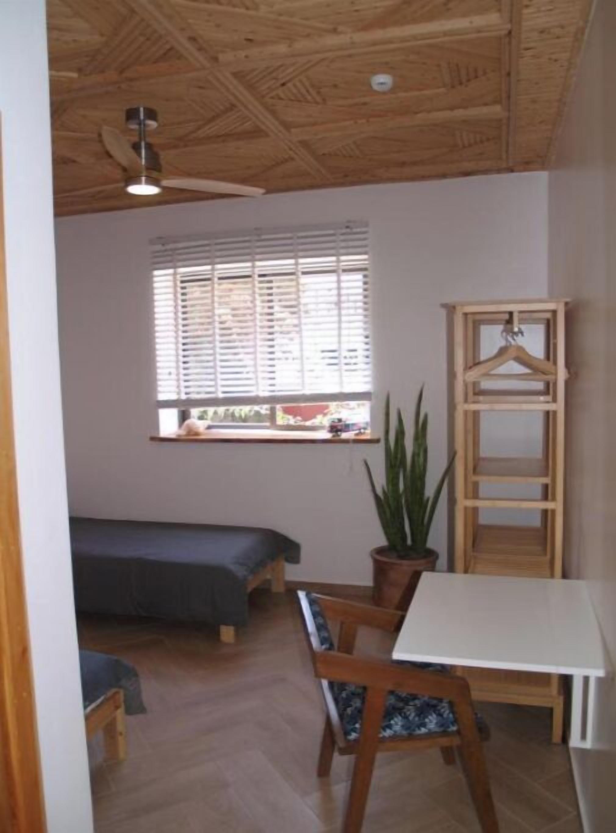 Quarto com teto de madeira, cama, mesa com cadeiras, prateleira suspensa e janela com persianas.