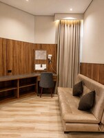 Quarto moderno com escrivaninha de madeira, cadeira cinza, sofá bege com almofadas e uma janela grande com cortinas.
