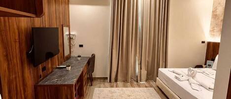 Quarto de hotel com uma secretária de madeira, uma cama, televisão e uma janela grande com cortinas.