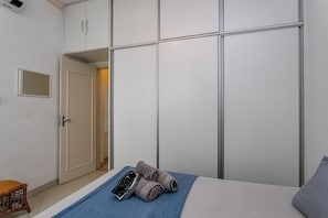 Um quarto com cama, uma toalha azul, uma toalha dobrada, um banquinho pequeno e um guarda-roupa com espelho.
