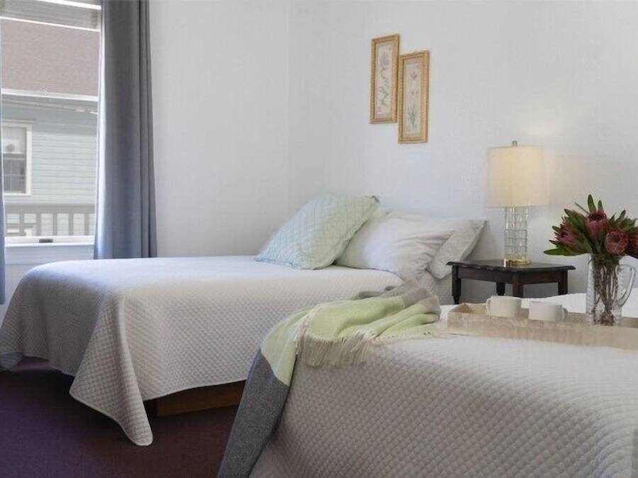 Quarto de hotel com duas camas, uma mesa de cabeceira com abajur, um vaso com flores e uma janela com cortinas.