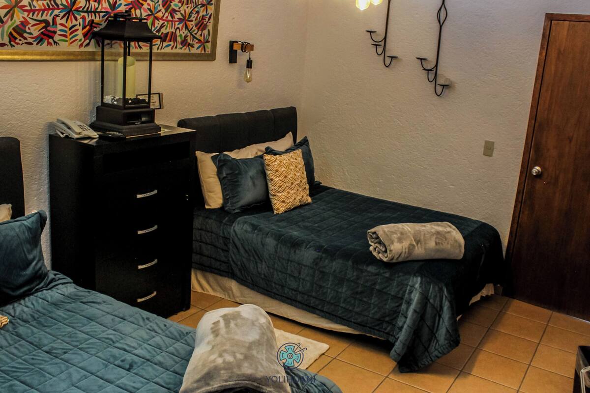 Quarto com duas camas, cômoda, abajur e luminária na parede.