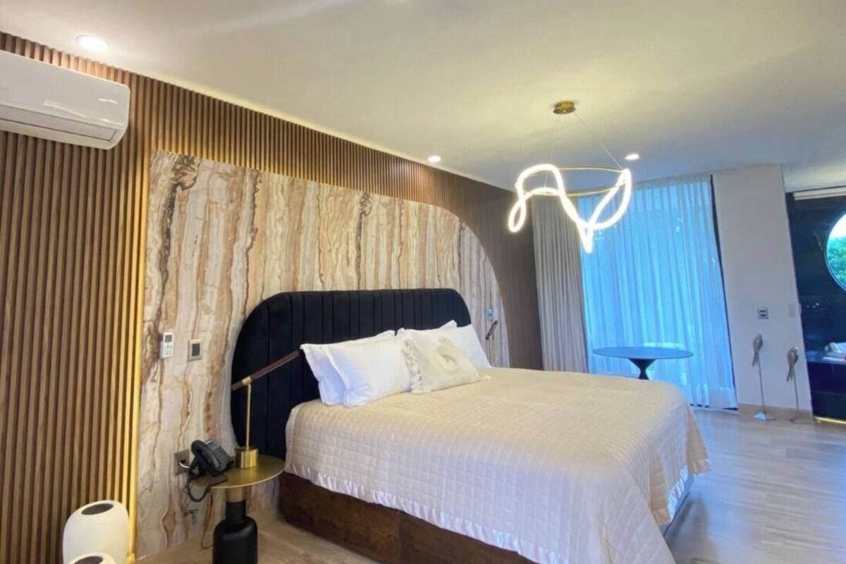 Quarto moderno com cama grande, lambris de madeira, ar condicionado e mesa redonda com luminária.