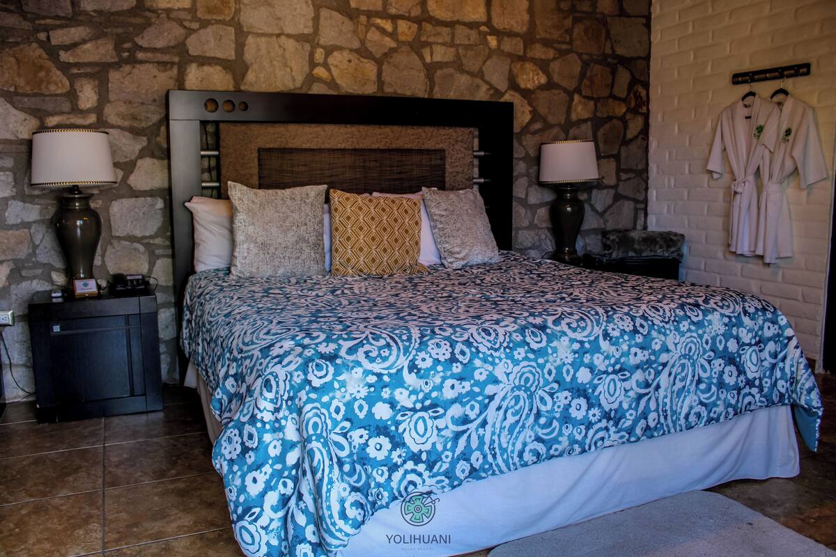 Quarto com parede de pedra, cama com roupa de cama estampada, duas mesas de cabeceira com luminárias e um porta-robes com roupões.