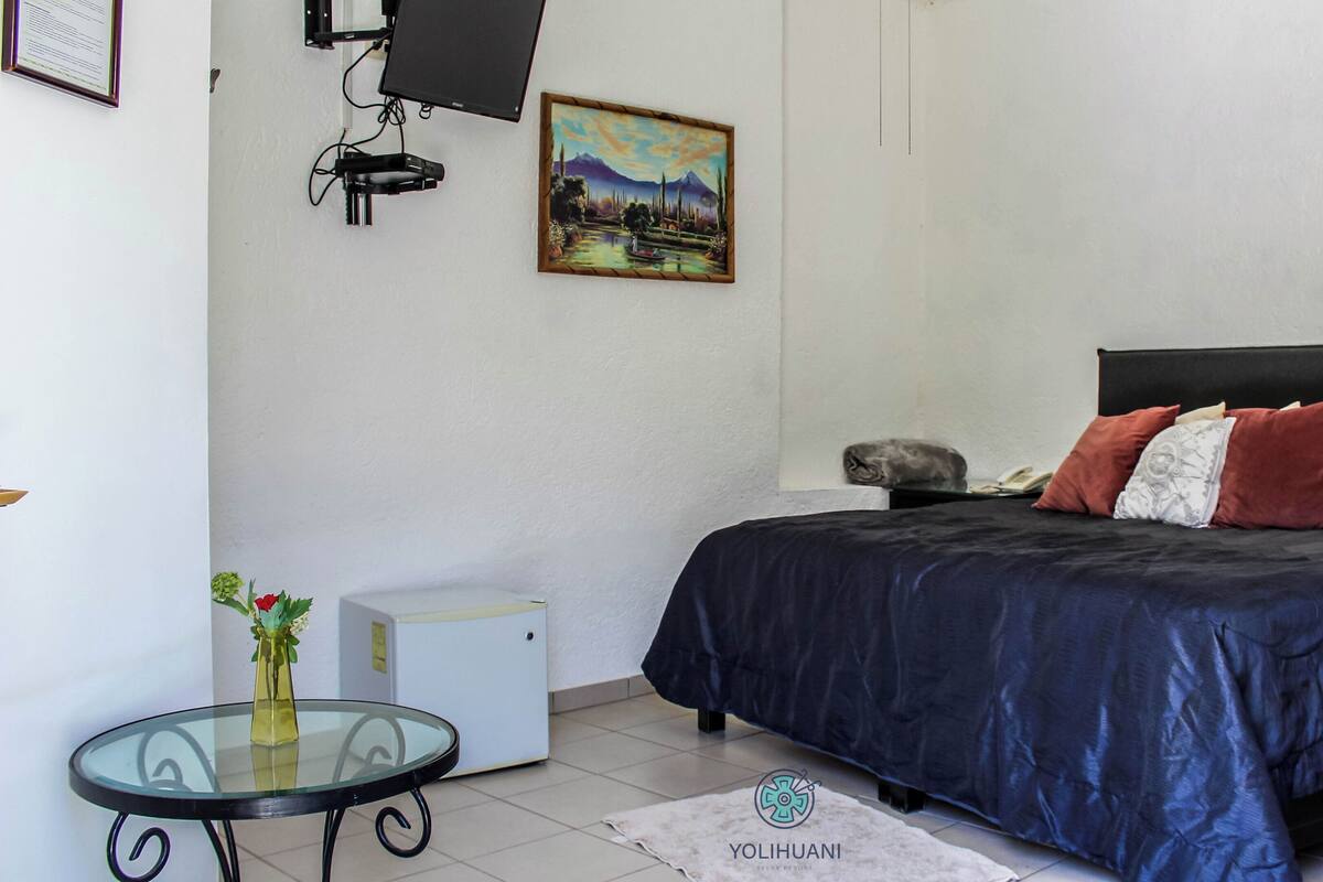 Quarto com cama, uma mesa pequena com um vaso, um projetor e um quadro na parede.