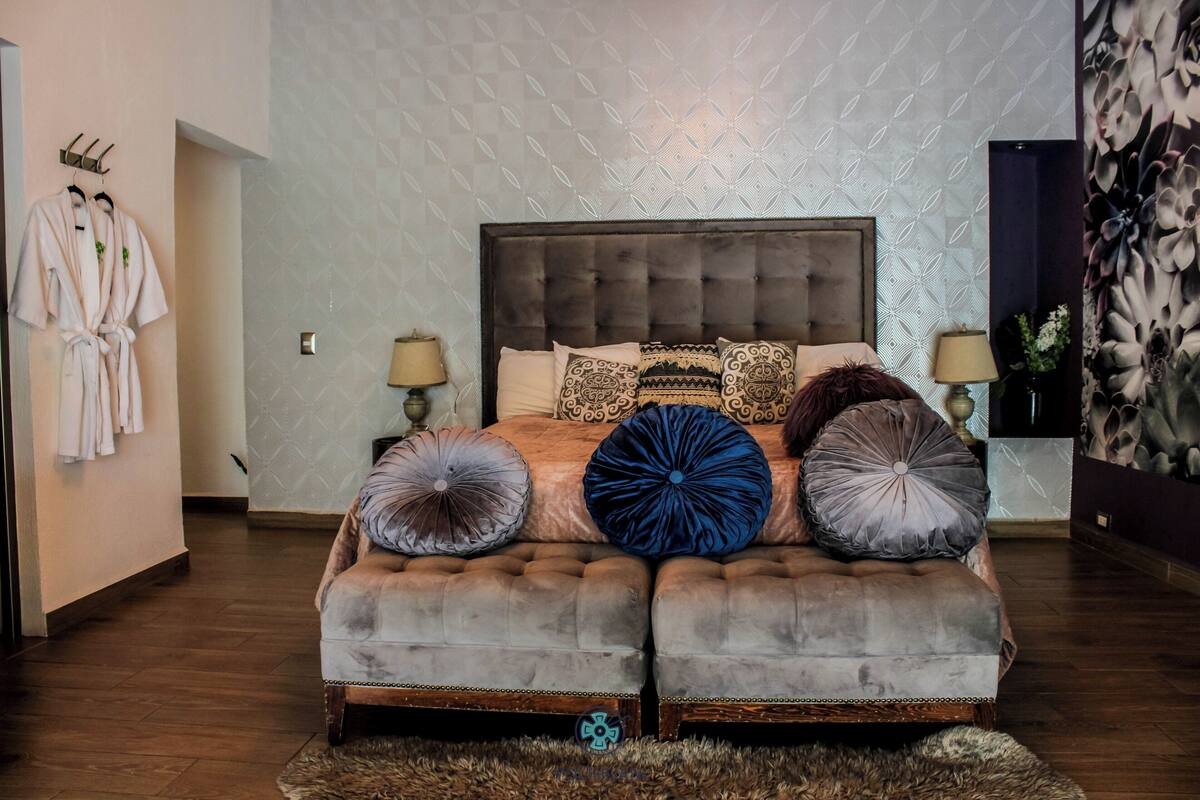 Quarto com cabeceira estofada, cama grande com várias almofadas, tapete fofo e parede com padrão geométrico.