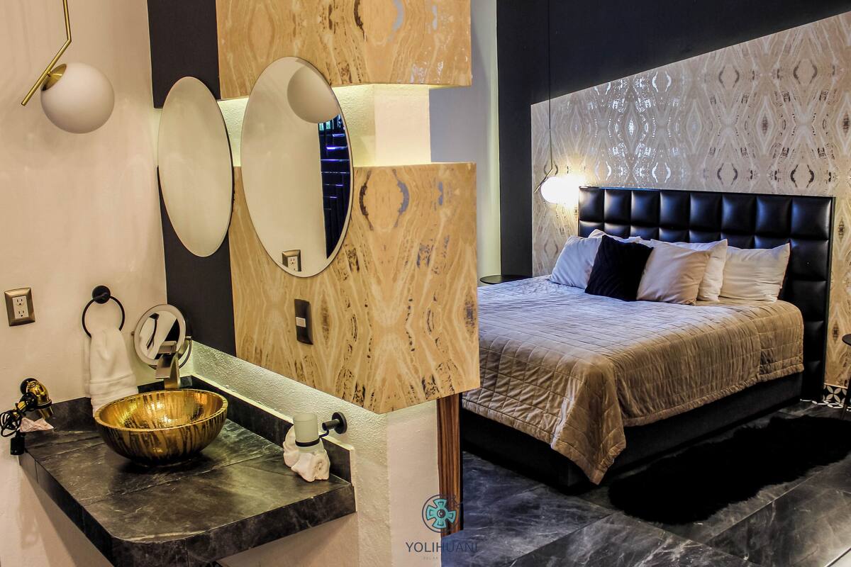 Casa de banho moderna com lavatório dourado, espelho e bancada preta. O quarto tem uma cama com cabeceira estampada, uma mesa de cabeceira e um criado-mudo.