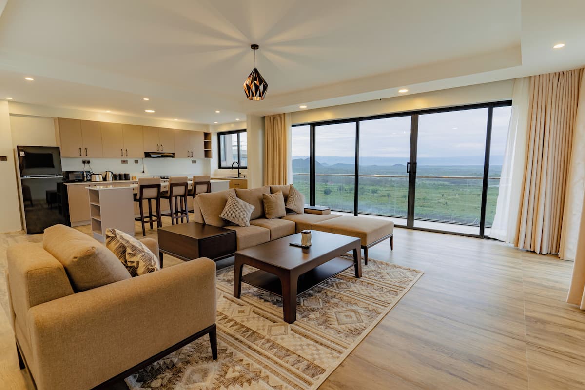 Uma sala de estar ampla, com sofá, mesa de centro e vista para uma paisagem verde através de grandes janelas.